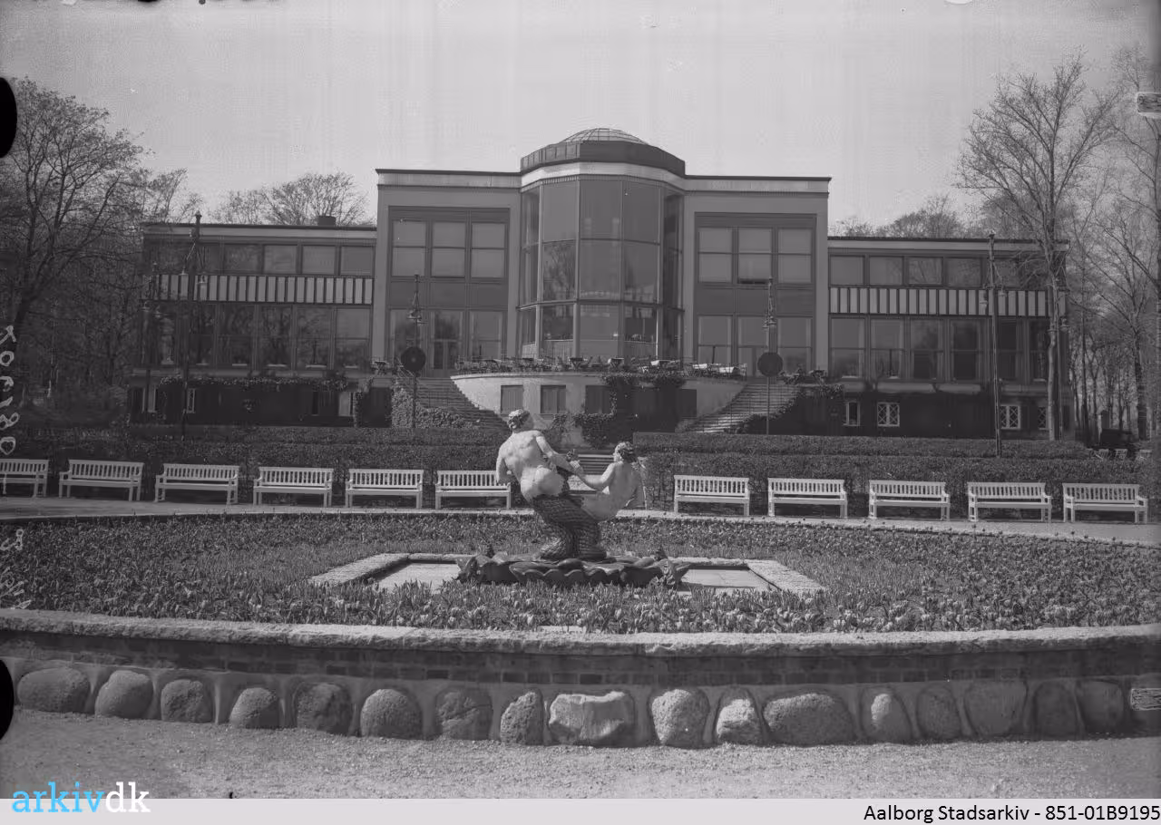 arkiv.dk | Restaurant Kilden set fra Kildeparken, med skulptur og bænke.